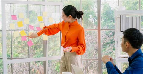 비즈니스 사람들은 사무실의 프로젝트 계획 보드에서 일하고 동료 친구와 대화하여 프로젝트 개발을 분석합니다 그들은 유리 벽에 게시 된 스티커 메모를 사용하여 정리합니다