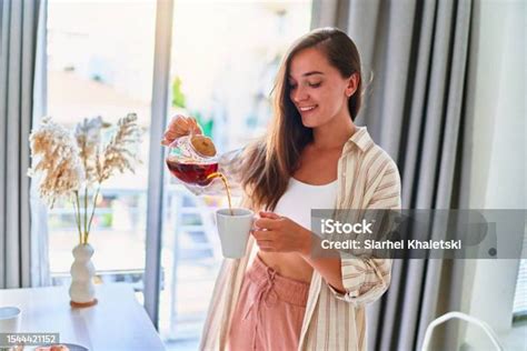 행복하고 귀여운 즐거운 미소를 짓는 만족스런 여자가 집에 있는 유리 주전자에서 컵에 뜨거운 차를 붓고 있다 차 뜨거운 음료에 대한 스톡 사진 및 기타 이미지 Istock