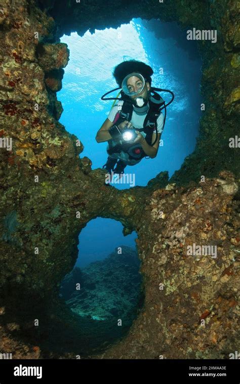 Female Diver Diving In Underwater Cave In Coral Reef Looking Through Opening Hole In Rock