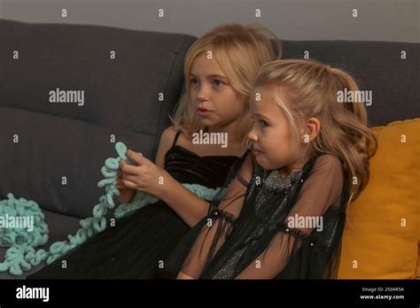 Two Blonde Girls In Black Dresses Sit On A Gray Couch One Holding A