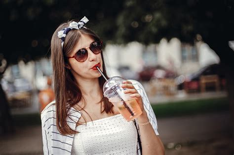 행복 한 Hipster 레모네이드를 마시는 밀 짚 플라스틱 컵을 들고과와 선글라스 레트로 드레스와 머리 띠 소녀 아름 다운 세련 된 젊은 여자 음료를 들고와 도시의 거리에서