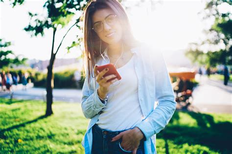 녹색 배경에 햇빛과 공원에서 야외서 스마트 폰에 연결된 Earnphones에서 재생 목록에서 전자 음악을 듣는 아름다운 쾌활한 젊은 여성의 절반 길이 초상화 갈색 머리에