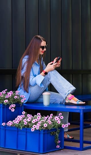 긴 금발 머리를 가진 소녀의 여름 초상화입니다 파란 셔츠에 선글라스를 낀 소녀가 카페 입구 벤치에 앉아 스마트폰으로 누군가와 대화를 나누고 있다 Brand Name