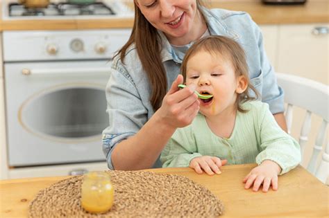 집에서 행복한 가족 부엌에 있는 숟가락으로 딸에게 먹이를 주는 어머니 지저분한 재미있는 얼굴을 가진 어린 아이는 집에서 건강에 좋은 음식을 먹는다 어린 딸에게 음식을 주는