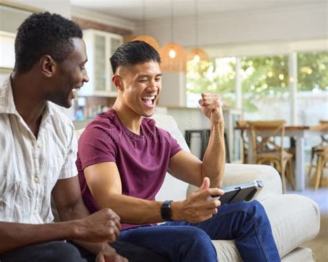 Same Sex Male Couple Or Friends On Sofa At Home With Handheld Mobile