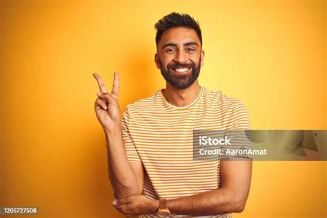 고립된 노란색 배경 위에 서있는 티셔츠를 입은 젊은 인도 남자가 손가락으로 승리 표지판을하고있는 카메라에 행복한 얼굴 윙크와 미소 2번 T 셔츠에 대한 스톡 사진 및 기타