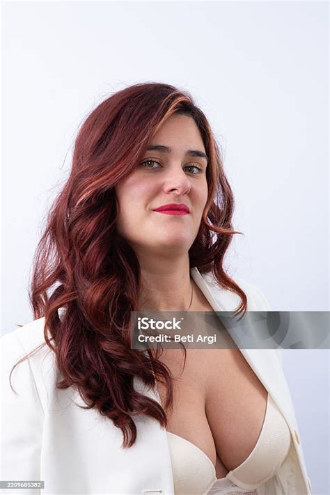 물결 모양의 빨간 머리와 빨간 입술을 가진 30 세 여성은 흰색 블레이저와 베이지 색 란제리를 입고 밝은 배경을 배경으로 자신감 있는 표정으로 웃고 있습니다 공간 복사