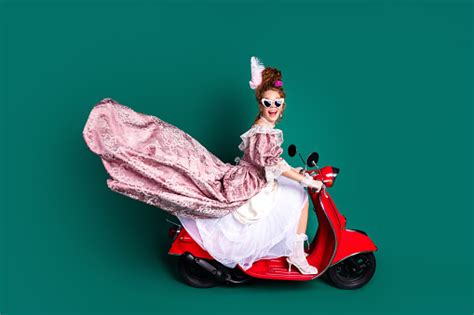 우아한 분홍색 바로크 양식의 드레스를 입은 여성이 녹색 배경의 빨간 스쿠터를 타고 역사와 현대 생활의 장난기 넘치는 혼합을 포착합니다 20 24세에 대한 스톡 사진 및 기타