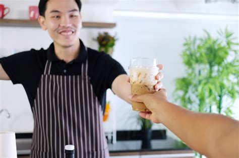 커피숍카운터바에서 아이스 라떼 커피 한 잔을 마시며 미소짓는 아시아 바리스타 청년 투명 플라스틱 유리를 들고 손에 초점을 닫습니다 2명에 대한 스톡 사진 및 기타 이미지