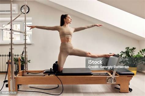 어린 소녀는 밝은 스튜디오의 리포머 침대에서 필라테스를 하고 있다 베이지색 바디수트를 입은 날씬한 갈색 머리는 균형을 훈련하고 다리 근육을 스트레칭하기 위해 운동을 합니다