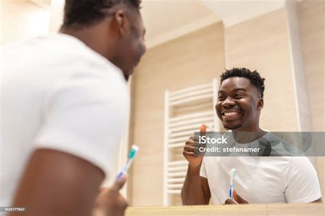 칫솔을 들고 미소를 짓고 욕실에서 거울을 보고 있는 아프리카 남자 남자는 화장실에서 아침에 자신의 이빨을 닦고 잠옷에 잠자는 남자가 잠들기 전에 밤에 치아를 닦아 20 24