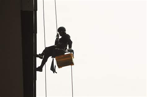 제복을 입은 산업 산악인 노동자와 헬멧 세척 파사드 교수형 벽의 실루엣 노동자 고층 로프 작업에 접근하십시오 산업 도시 작품