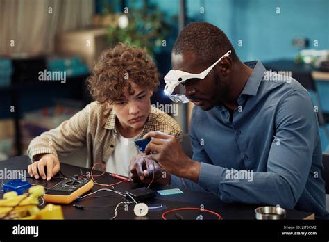 Portrait Of Young Teenage Boy Building Robots In Engineering Class With Male Teacher Helping