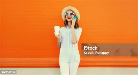 오렌지색 배경에 밀짚모자를 쓰고 커피 한 잔을 들고 스마트폰을 부르는 행복한 웃는 젊은 여성 20 29세에 대한 스톡 사진 및 기타 이미지 Istock