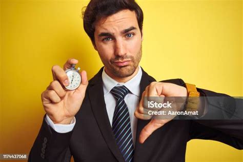 화가 얼굴 엄지 손가락으로 싫어하는 것을 보여주는 부정적인 기호 거부 개념으로 고립 된 노란색 배경 위에 서있는 스톱워치를 들고있는 젊은 잘생긴 사업가 실패에 대한 스톡