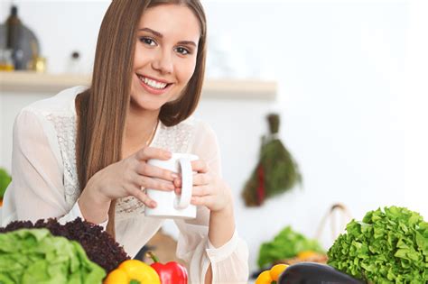 젊은 행복 한 여자 흰색 컵을 들고와 녹색 채소 중 부엌에 있는 나무 테이블에 앉아있는 동안 카메라를 보고 가정 생활에 대한 스톡 사진 및 기타 이미지 Istock