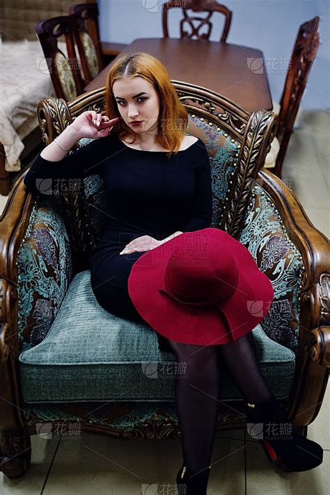 时尚的红发女孩，戴着红色的帽子，穿着黑色的礼服，浓妆艳抹，坐在皇家扶手椅上。