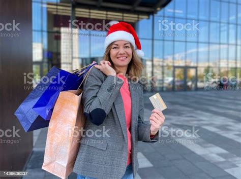 백인 쾌활한 여성 산타 모자를 머리에 쓰고 많은 쇼핑백과 신용 카드를 들고 도시의 야외에 서 있는 카메라에 미소를 짓고 있다 크리스마스 선물 구매 40 44세에 대한 스톡