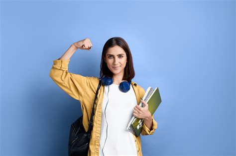 행운을 빌어 강한 웃는 학생 소녀는 근육을 보여주고 그녀의 지식에 자신있게 이정표를 통과하고 책을 들고 노란색 셔츠 흰색 티셔츠 검은 색 가방과 헤드폰을 목에 걸고 있습니다