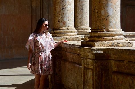 밝은 분홍색 여름 드레스와 선글라스를 쓴 젊은 갈색 머리 여자는 돌 주랑으로 둘러싸인 벽에 기대어 앞을 내다보고 있다 스페인