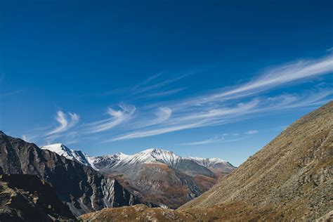 푸른 하늘의 서러스 구름 아래 햇빛에 눈이 와 가을 색상의 큰 산과 다채로운 고산 풍경 햇살이 내리쬐는 바위와 눈 덮인 산 꼭대기가 있는 그림 같은 가을 풍경 가리기에 대한