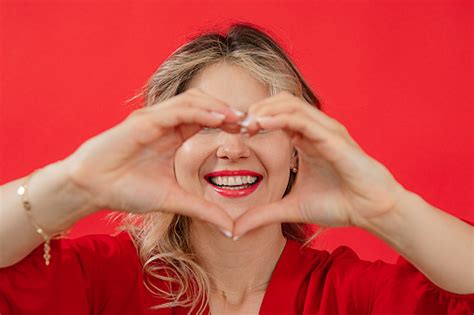 빨간 배경에 격리된 완벽한 밝은 화장을 한 빨간 옷을 입고 하얀 눈 미소를 짓고 있는 행복한 여자의 초상화 근접 촬영 얼굴 높이에서 손으로 하트 모양을 만드는 사랑 표현