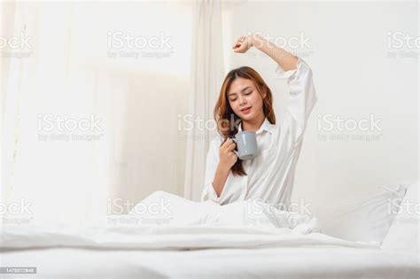 하얀 잠옷을 입은 여자가 주말 아침에 일어나 휴식을 취하고 노트북 휴대폰을 가지고 놀며 하얀 침실 안의 유리에서 빵을 먹고 차를 마신다 아침 휴가 개념입니다 갈색 머리에