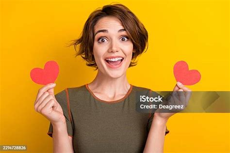 짧은 머리를 한 놀란 기쁨에 넘친 소녀의 초상화는 카키색 티셔츠를 입고 노란색 배경에 고립 된 두 개의 종이 심장을 들고 있습니다 Omg에 대한 스톡 사진 및 기타 이미지