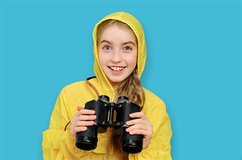 노란 재킷을 입은 백인의 즐거운 소녀는 쌍안경을 손에 들고 있다 하이킹 아이디어와 캠핑 장비 스튜디오 는 파란색 배경에 반 길이의 초상화를 분리 10 11세에 대한 스톡