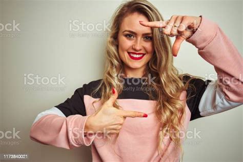 행복한 얼굴로 손과 손가락으로 프레임을 만드는 흰색 고립 된 배경 위에 서있는 스웨트 셔츠를 입은 젊은 매력적인 여성 창의력과