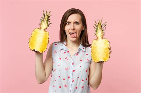 분홍색 파스텔 배경에 고립 된 신선한 잘 익은 파인애플 과일의 Halfs를 들고 혀를 보여주는 여름 옷에 젊은 여자를 의아해 사람들은 생생한 라이프 스타일 휴식 휴가