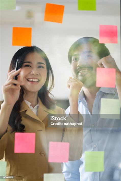 웃고 있는 창의적인 사업가들은 사무실의 유리 벽에 붙은 스티커 메모를 보고 브레인스토밍을 하고 있다 가리키기에 대한 스톡 사진