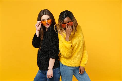 두 젊은 여자 친구는 밝은 오렌지 벽 배경 스튜디오 초상화에 고립 된 포즈를 서 있는 검은 노란 옷에 유럽과 아프리카계 미국인 사람들 라이프 스타일 개념입니다 복사 공간을