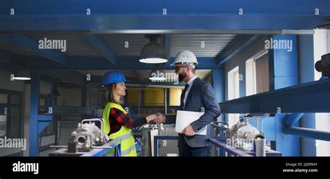 Engineer And Industrial Worker In Uniform Shaking Hands In Large Metal Factory Hall And Talking