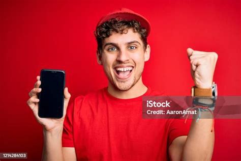 젊은 금발 배달 남자 와 곱슬 머리 입고 모자 를 들고 스마트 폰 을 들고 스마트 폰 위에 자랑스럽게 비명을 지르며 승리와 성공을 축하하는 매우 흥분 응원 감정 감정에 대한