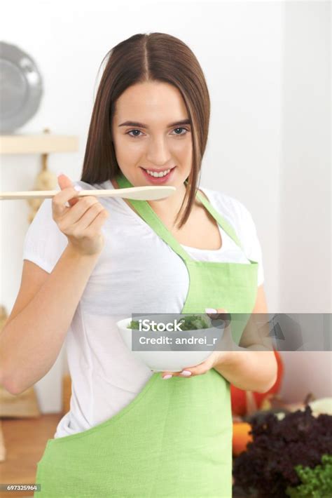 녹색 앞치마에 젊은 갈색 머리 여자 요리 또는 부엌에서 신선한 샐러드를 먹는 나무 숟가락과 그릇에 그녀의 손 잡고 주부 음식과