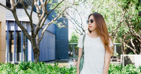 흰색 빈 티셔츠를 입은 아시아 힙스터 소녀 긴 갈색 머리가 길 한가운데에 서 있다 스트리트웨어를 입은 여성이 녹색 도시 배경에