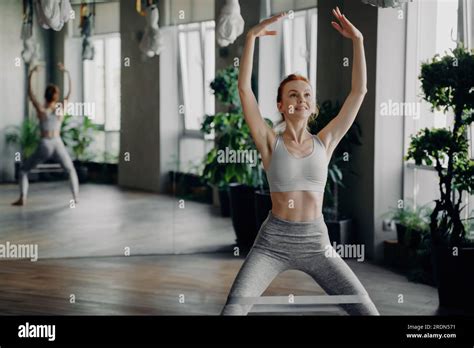Slender Redhead Fitness Instructor Enjoys Squats With Pilates Resistance Band In Gym Hands