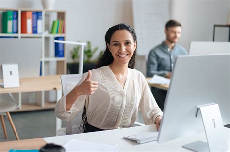 현대 사무실에서 컴퓨터로 작업하는 동안 엄지손가락을 치켜드는 제스처를 보여주는 쾌활한 젊은 여성 직원 엄지손가락을 위로에 대한 스톡 사진 및 기타 이미지 Istock