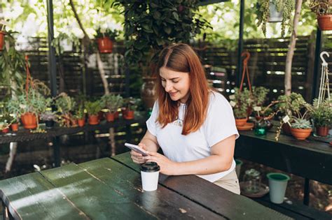 커피 종이 컵을 들고 카페에 앉아있는 동안 스마트 폰을 보는 젊은 아름다운 여성 휴대 전화를 사용하여 커피와 함께 테이블에 앉아있는 행복한 소녀 여름 시간 30 39세에