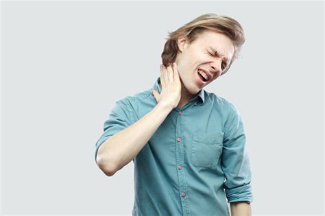 목 통증 아픈 잘생긴 긴 머리 금발 젊은 남자의 초상화는 나쁜 느낌 때문에 그의 고통 스러운 목을 들고 서 있는 파란 캐주얼 셔츠 가꿔주기에 대한 스톡 사진 및 기타 이미지