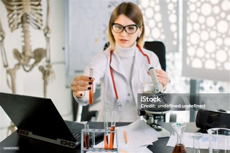 실험실에서 노트북 현미경으로 테이블에 앉아있는 동안 그녀의 손에 빨간 액체와 과학 샘플 테스트 튜브를보고 젊은 예쁜 백인 여성 과학자 손에 초점 Stem 주제에 대한 스톡