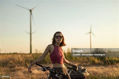 해질녘 야외에서 산악 자전거를 탄 아름다운 여성의 여성 초상화 아름다운 사람들과 함께하는 라이프스타일 사진 선글라스를 쓴 사람 풍력 발전 단지의 터빈 녹색 에너지 기술로