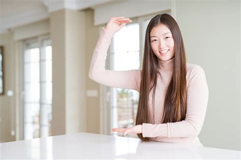 크고 큰 크기의 기호를 보여주는 손으로 흰색 테이블에 캐주얼 스웨터를 입고 아름 다운 아시아 여자 기호를 측정 합니다 카메라를 바라보며 웃고 있습니다 측정 개념 가정 생활에