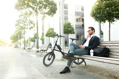 재사용 가능한 커피 컵에서 전기 자전거 음료 커피를 마시는 비즈니스 통근자 및 팟 캐스트를 듣습니다 도시에 대한 스톡 사진 및