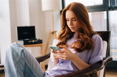 Portrait Of Thoughtful Focused Young Woman Texting Message On Mobile Phone Sitting On Arm Chair