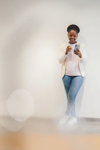 흰 벽 배경에 서서 웃고 있는 아프리카 사업가의 전체 길이 초상화 커피 한 잔을 들고 휴대 전화를 사용합니다 통신 네트워크 흰색 배경입니다 25 29세에 대한 스톡 사진 및