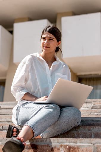 하얀 셔츠와 청바지를 입은 젊은 검은 머리 여성은 여름날 도시 거리의 계단에 앉아있는 노트북을 사용합니다 원격 근무 프리랜서