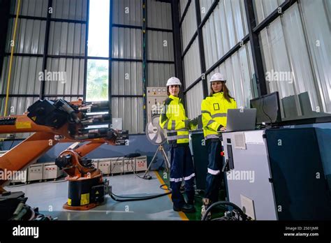 Two Engineers In Safety Gear Stand Beside A Robotic Arm Inside A Bright Industrial Space They