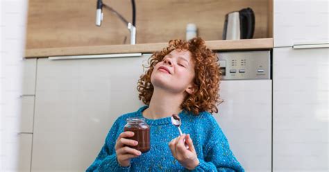 나무 부엌 바닥에 앉아 항아리에서 초콜릿을 먹는 젊은 여자 숟가락을 사용하여 항아리에서 초콜릿을 먹고 한 입 가득 맛보는 건방진 얼굴에 탐닉하는 귀여운 생강 소녀 사진